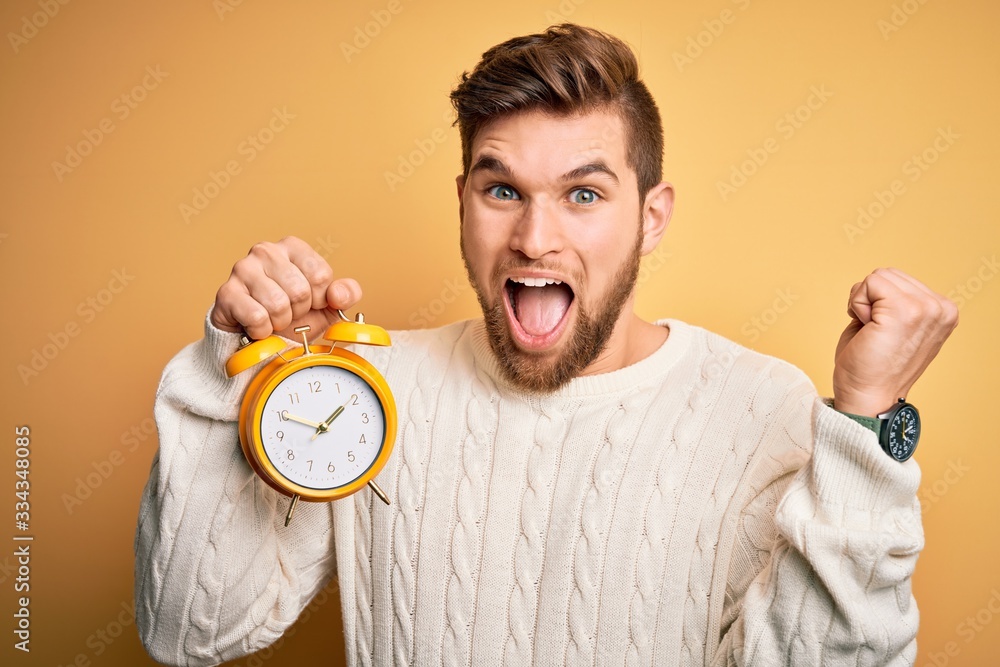 Young blond man with beard and blue eyes holding alarm clock over ...