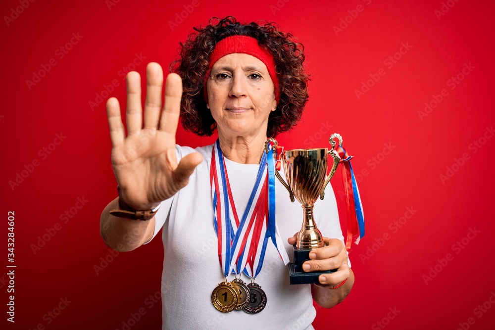 Middle age curly woman winning medals holding trophy over isolated red ...