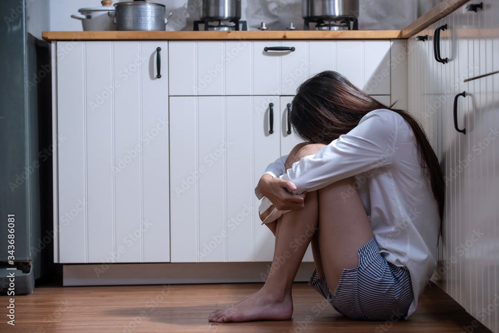 Depressed young beautiful Asian woman sitting on kitchen floor with hugging knees. Loneliness ...