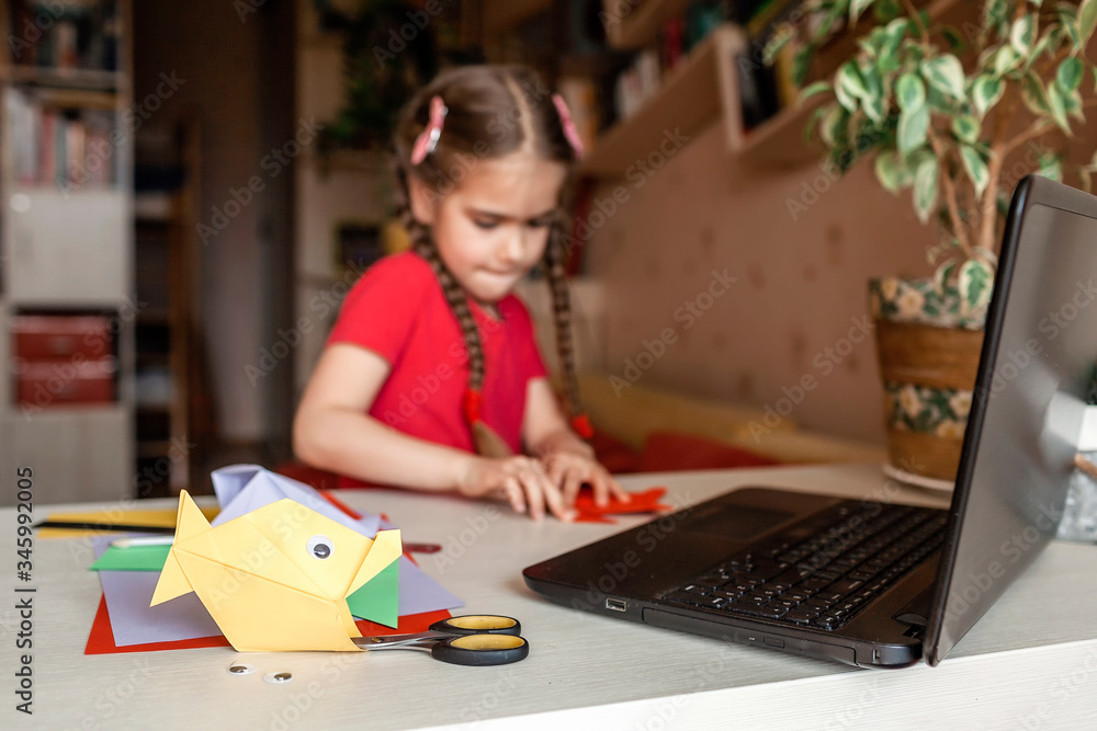 Girl doing origami fish with color paper looking video on laptop ...