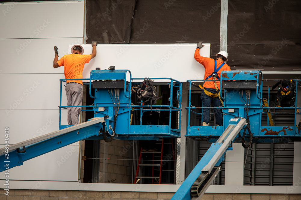 Two construction workers wearing hardhats on a blue manlift hanging ...