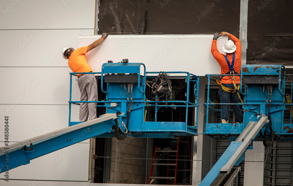 Two construction workers wearing hardhats on a blue manlift hanging ...