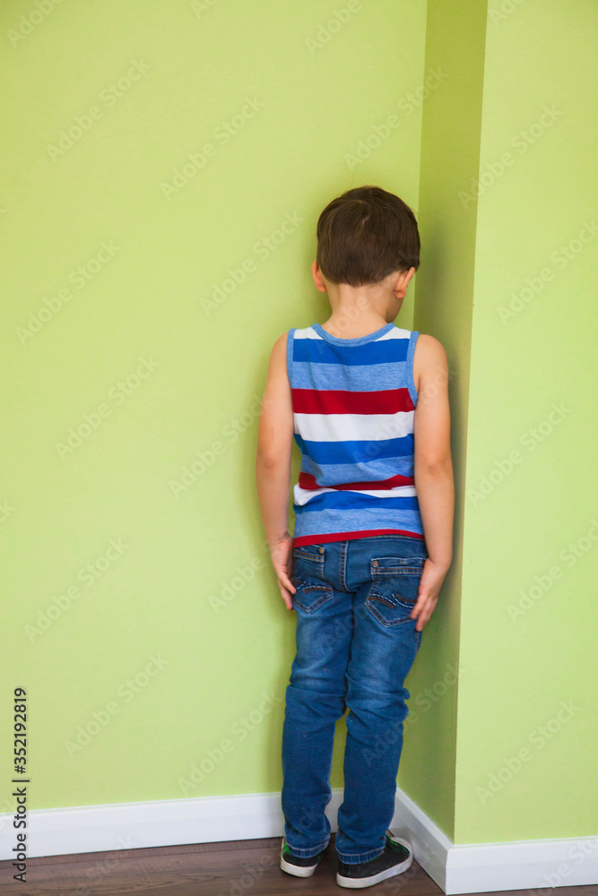 a young boy in a striped t-shirt stands in the corner with his face ...