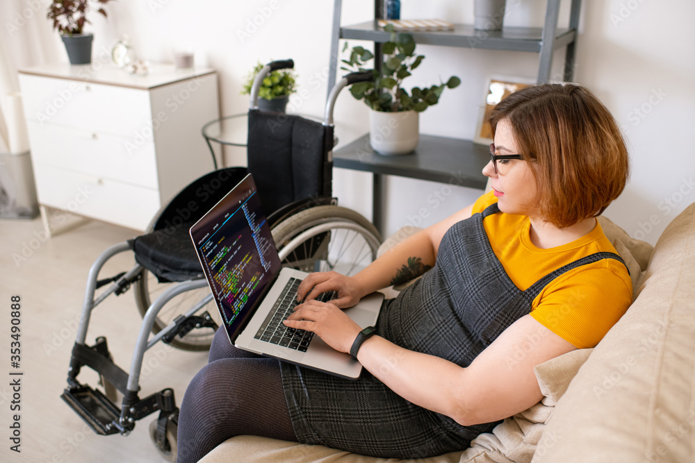 Serious disabled woman in eyeglasses sitting on sofa and using laptop ...