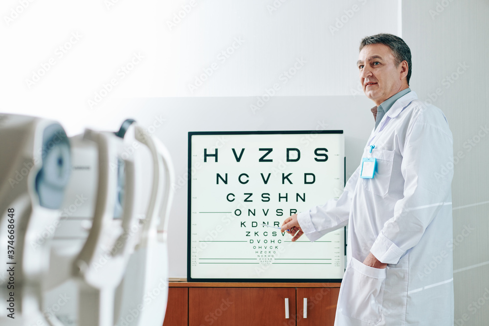 Mature ophthalmologist pointing at letters on test chart when checking ...