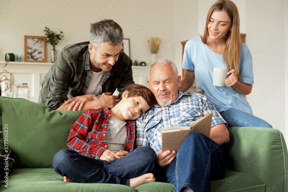 Grandfather reading book aloud to cute schoolboy and his smiling family. Multi-generational ...