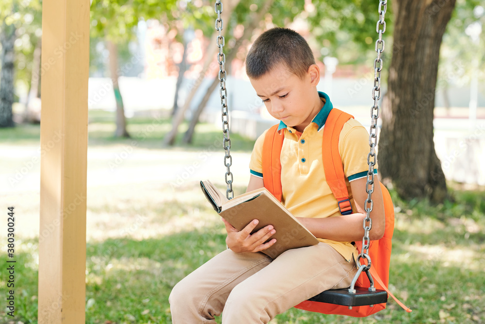 Curious boy with satchel on back sitting on swing in public park and ...