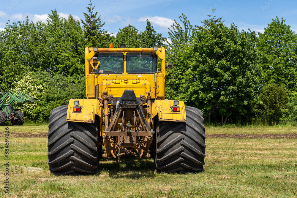 Backside of a yellow tractor with heavy wheels in a field with trees in ...