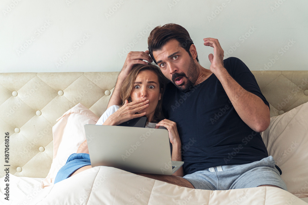Scared young couple sitting under blanket on the bed with laptop while ...