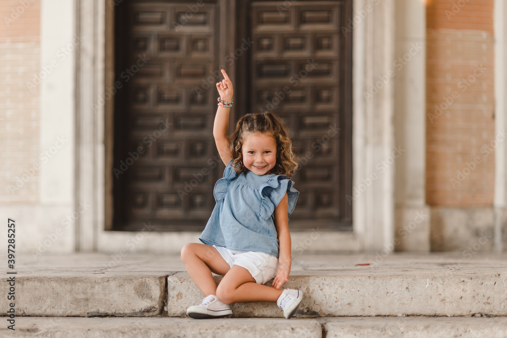 Positive little child in summer outfit sitting with raised arm on ...