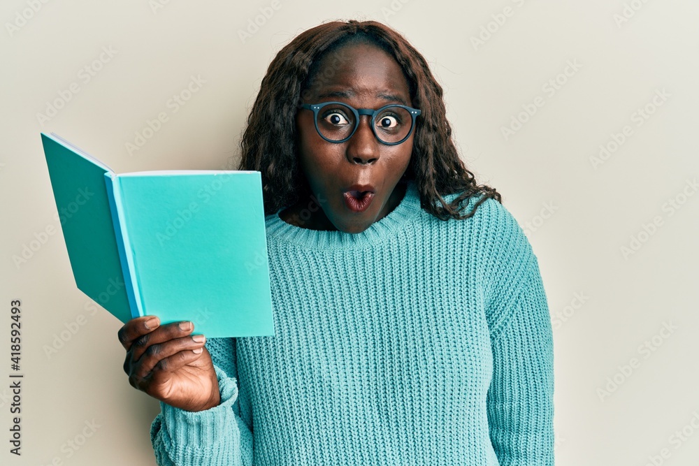 African young woman reading a book wearing glasses scared and amazed ...