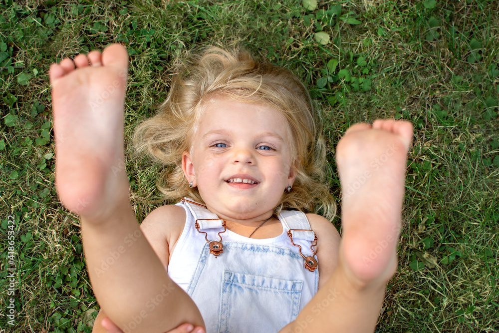 Happy blond girl lying on the green grass raising her legs up and ...