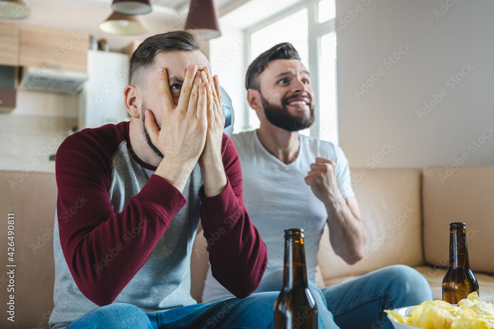 Nervous concentrated men sitting on sofa watching soccer play live broadcast on tv and cheering ...