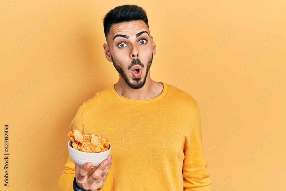 Young hispanic man with beard holding nachos potato chips scared and ...