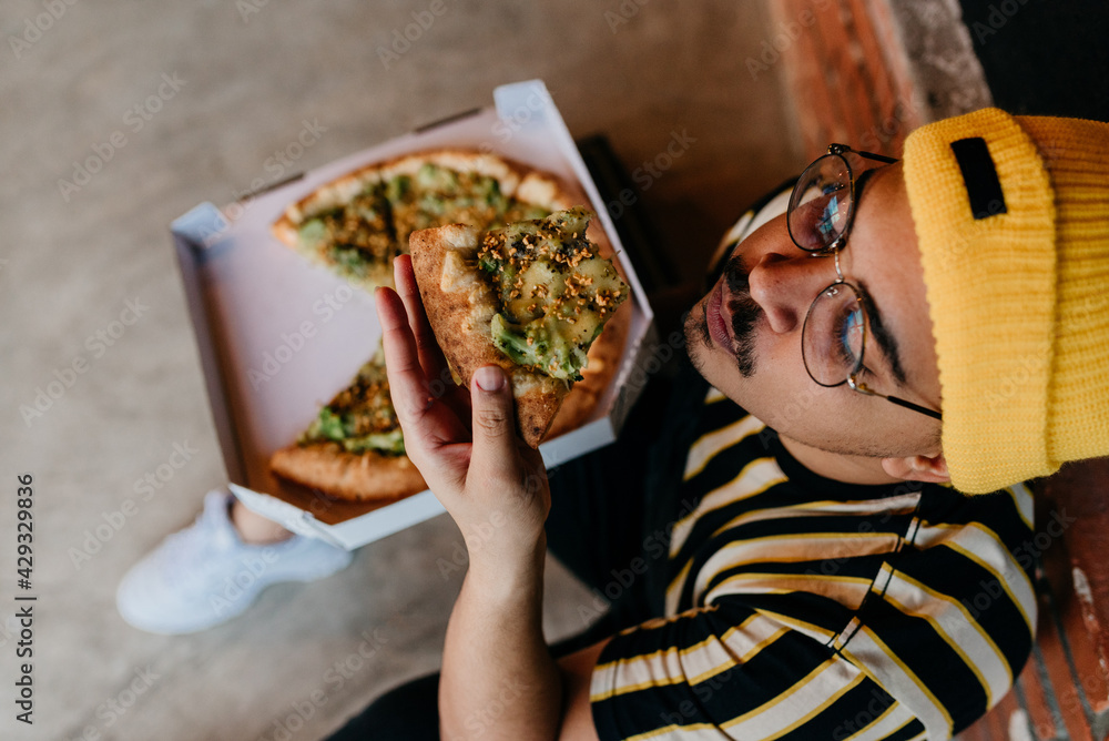 male model sitting on the floor eating a piece of pizza with his hand ...