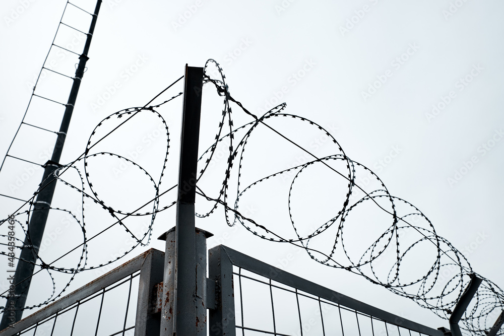 Barbed wire over a metal wall with a grid and a tower with gray sky ...