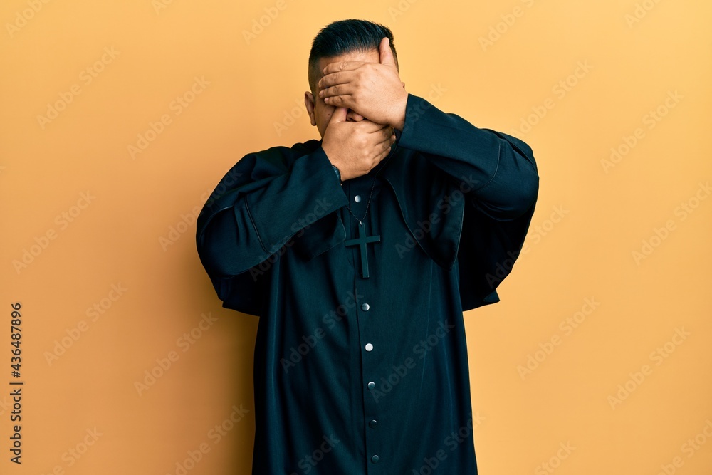 Young latin priest man standing over yellow background covering eyes ...