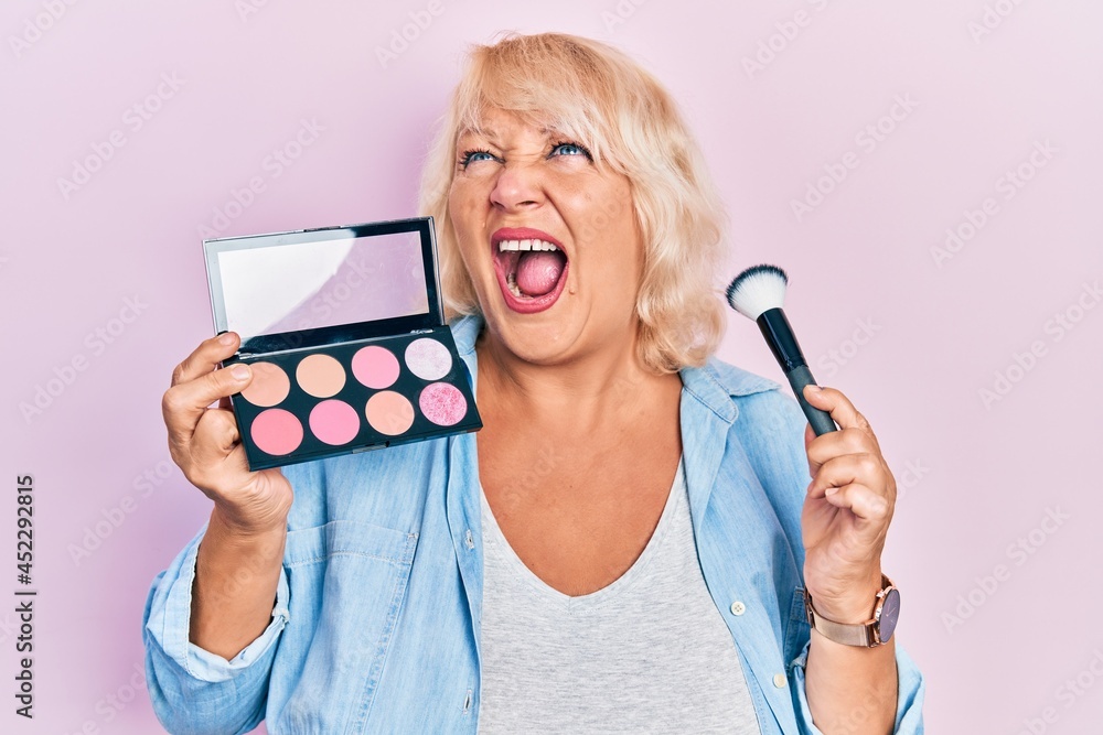 Middle age blonde woman holding makeup brush and blush angry and mad screaming frustrated and ...