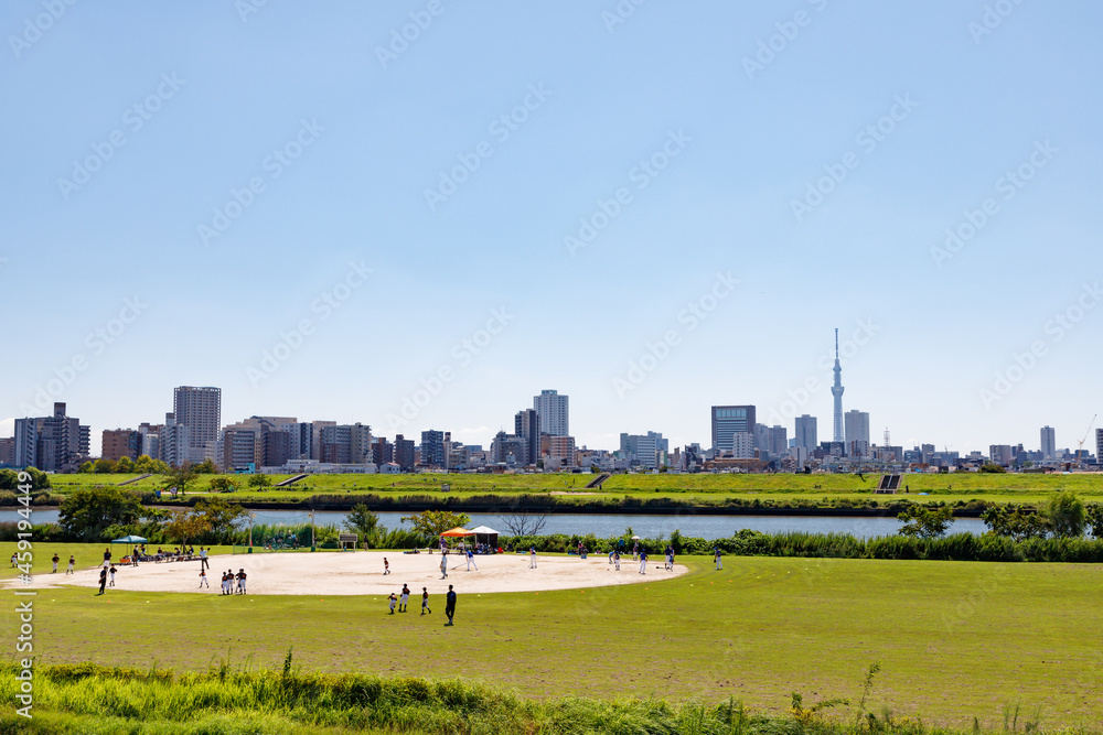Стокове фото 青空を背景に足立区側から見た荒川区方面の景色と河原のグランドで野球をする人々 Adobe Stock