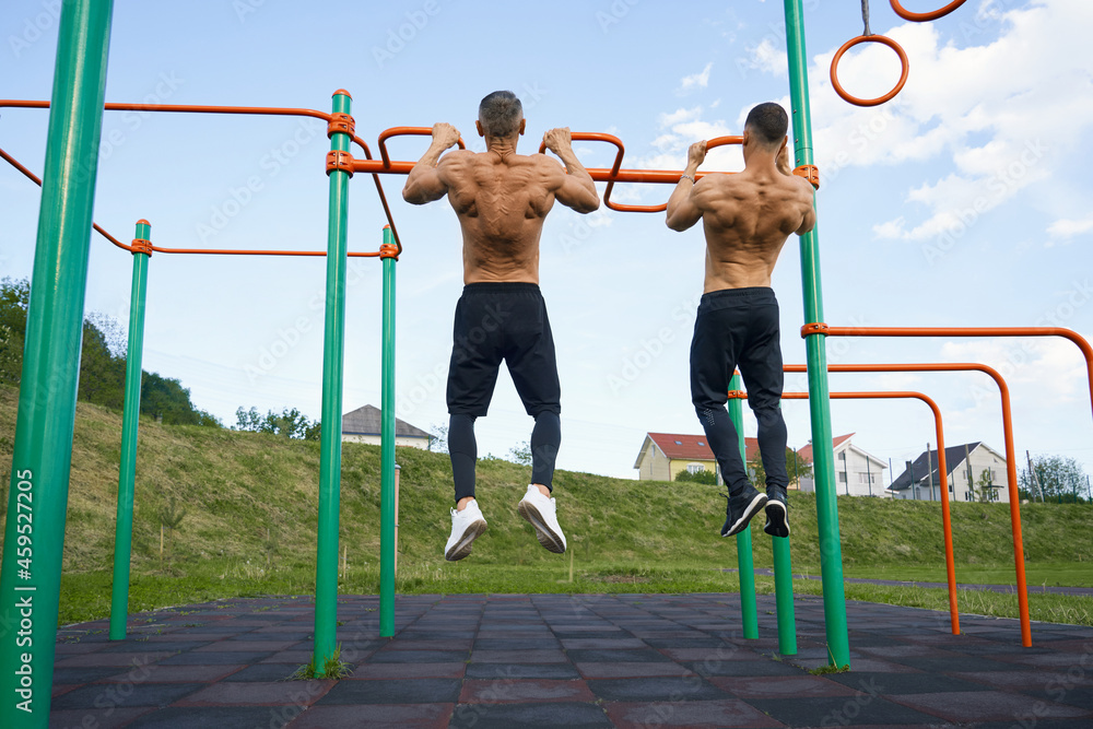 Back view of two shirtless bodybuilders doing pull ups on sports ground ...