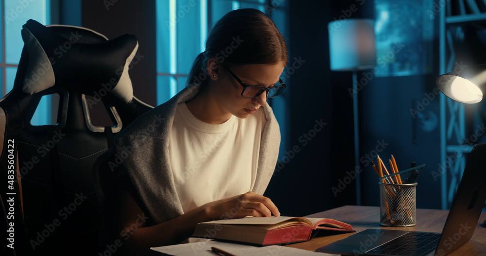 Smart schoolgirl student in eyeglasses reading book late at night ...