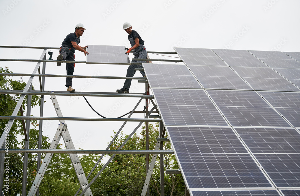 Workers installing solar panels on metal beams. Men wear helmets and ...