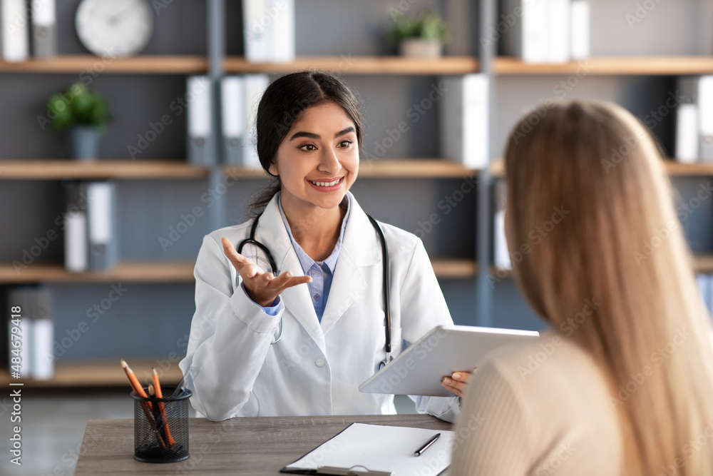 Smiling young attractive indian lady medical worker in white coat gestures and consultation ...