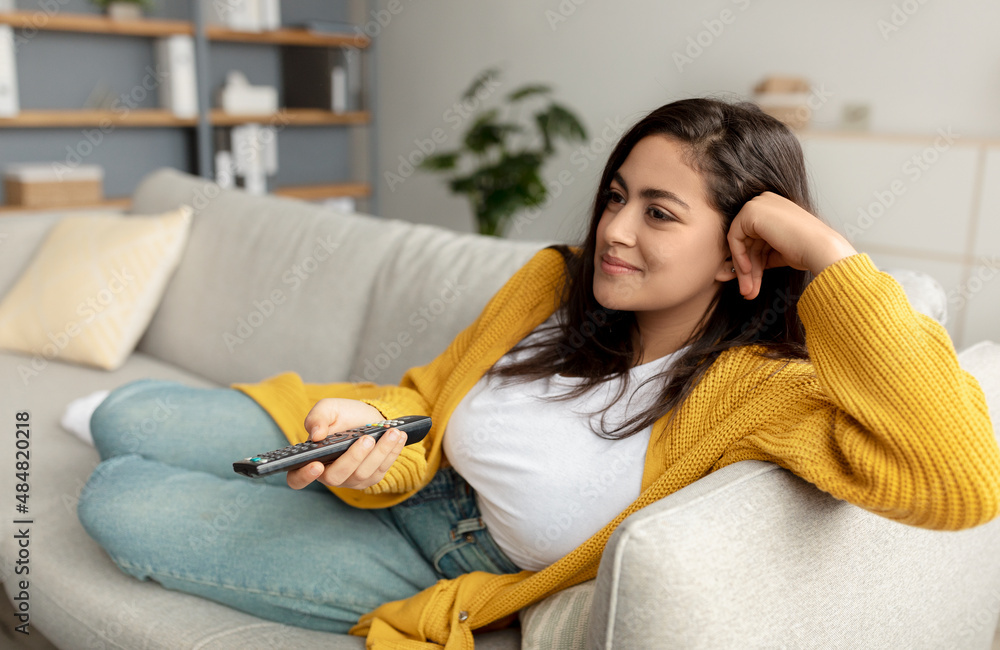 Pretty arab lady with remote control watching TV on couch at home ...