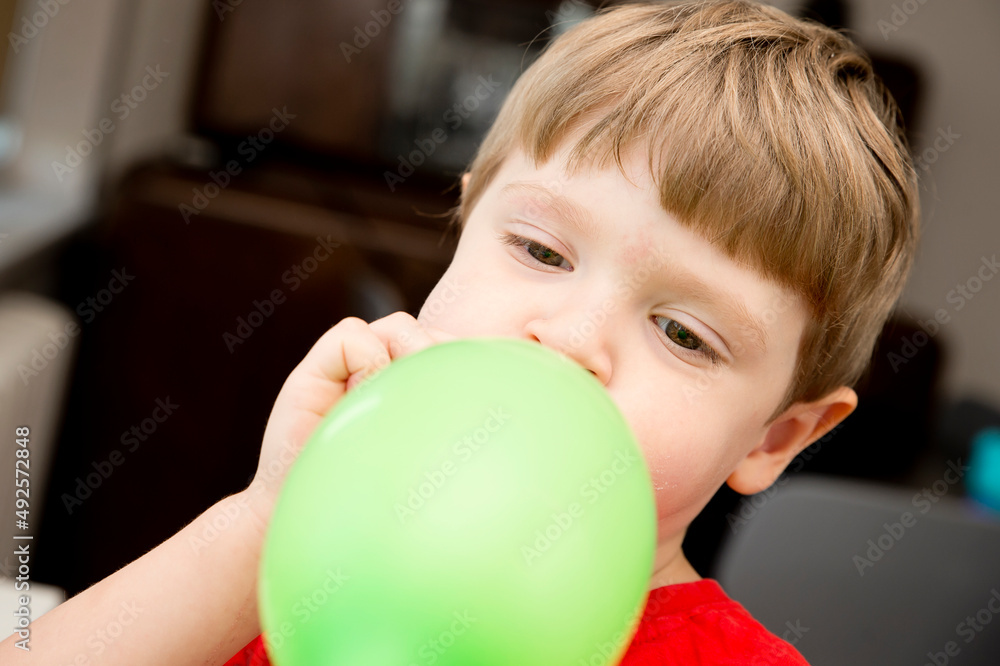 3 year old boy inflating green balloon with a mouth. Swollen cheeks and ...