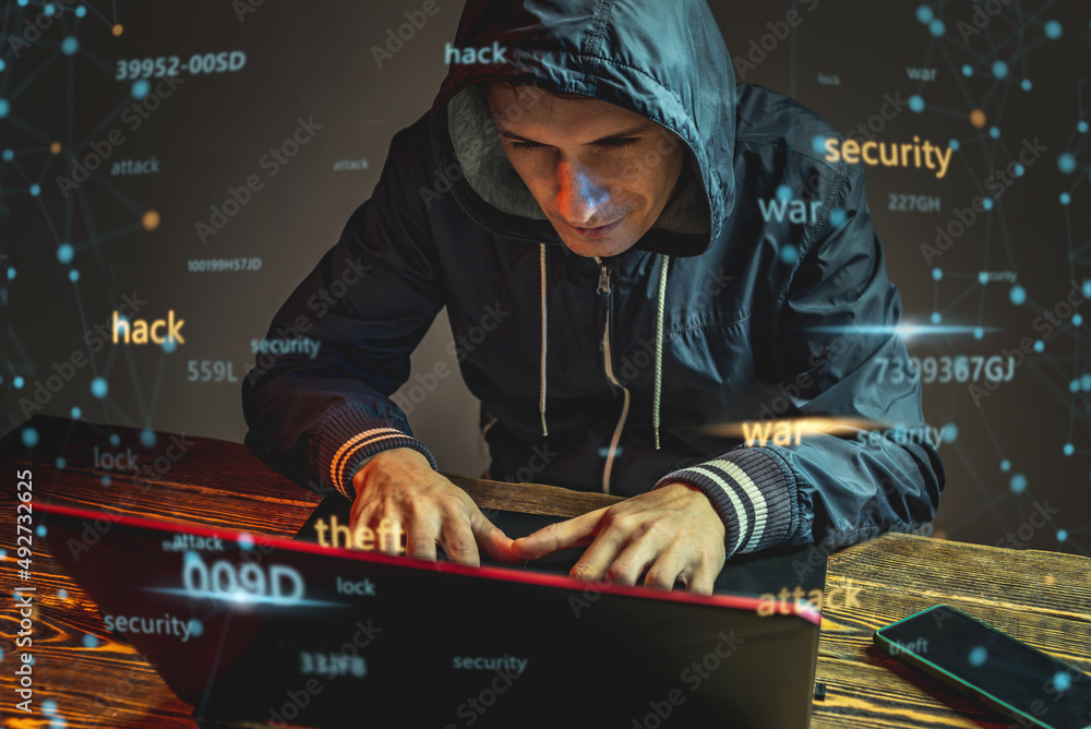 Hacker in a hood with a phone is typing on a laptop keyboard in a dark room. Concept of cyber ...