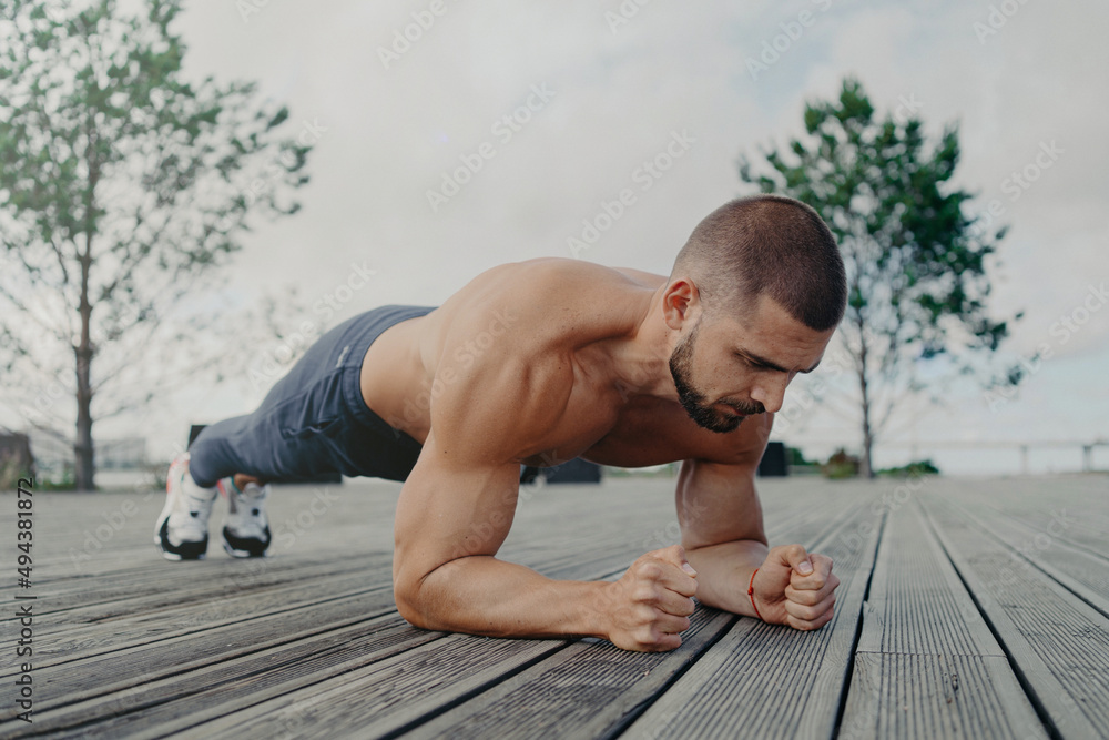 Outdoor shot of strong sportsman stands in plank pose, puts all efforts ...
