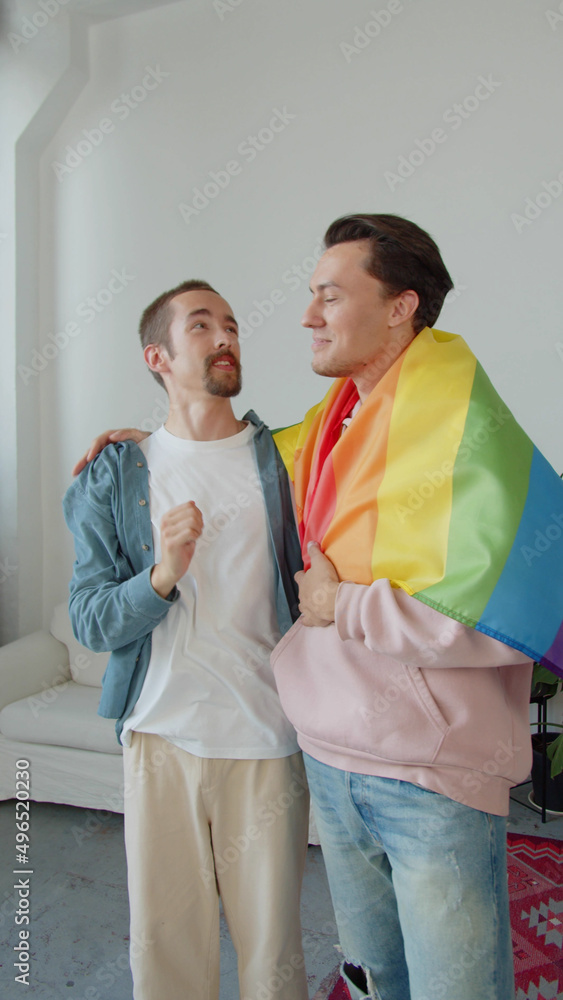 Two young men stand together, covered with a lgbt flag, and one of them ...