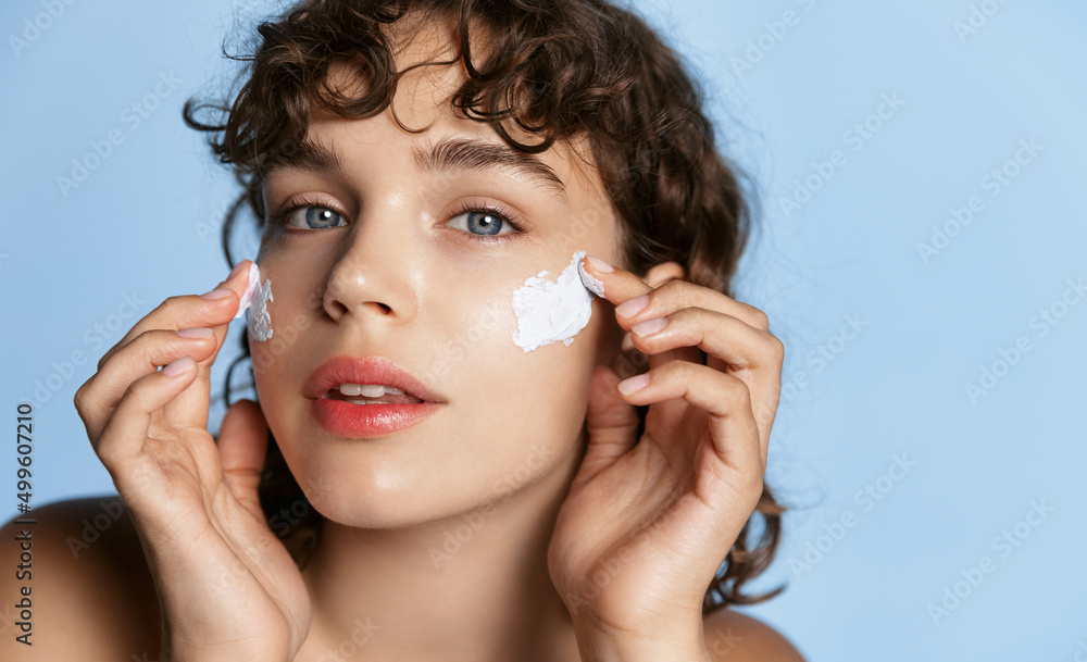 Young woman looking in mirror and applying facial cream on her cheeks ...