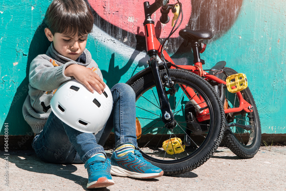 sad and angry little boy with bycicle and helmet sitted on the ground ...