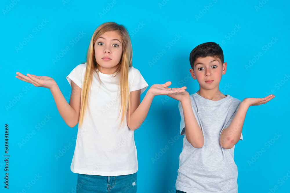 Puzzled and clueless two kids boy and girl standing over blue studio ...