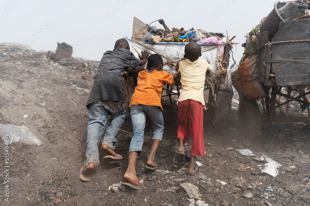Three black street boys pushing a heavy garbage cart up a heap in urban ...