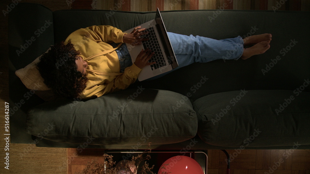 One young black girl lying down on couch opening laptop. Top view angle ...