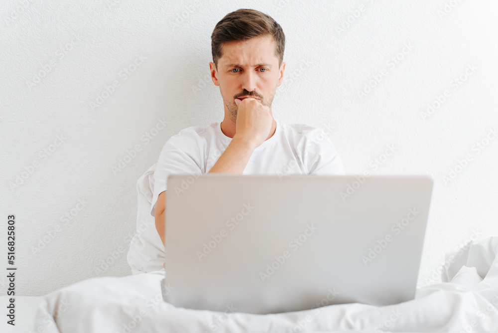 Thoughtful busy young man using laptop while sitting on bed in bedroom ...