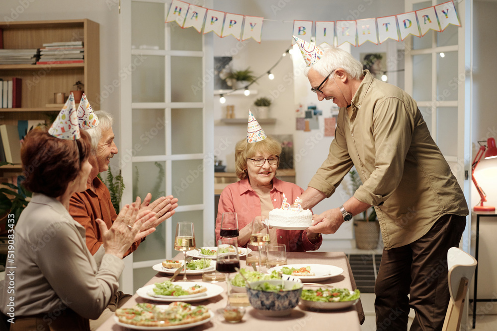 Cheerful aged man in cap putting delicious birthday cake on served ...