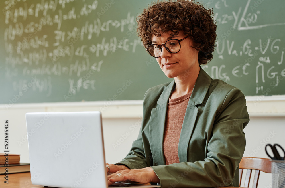 Young confident teacher typing on laptop keypad while sitting by ...