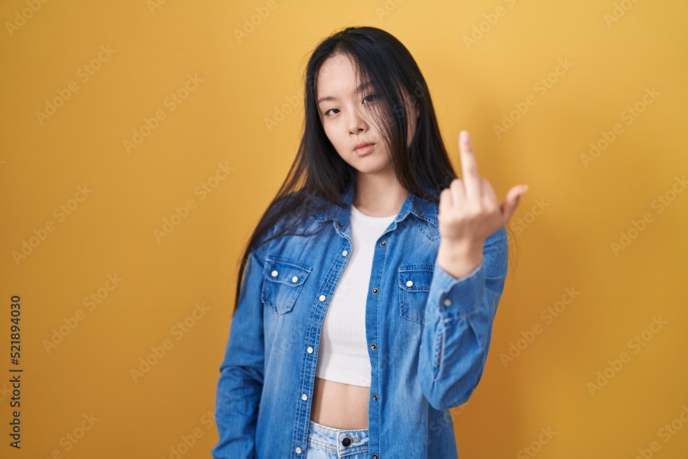 Young asian woman standing over yellow background showing middle finger ...