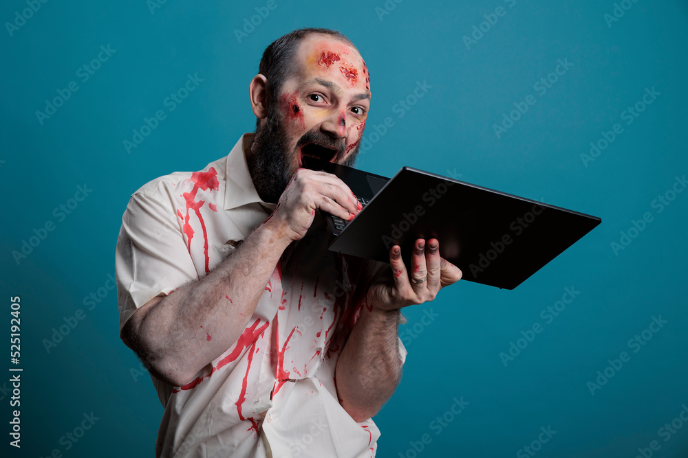 Scary zombie biting on laptop computer, having bloody scars and wounds ...