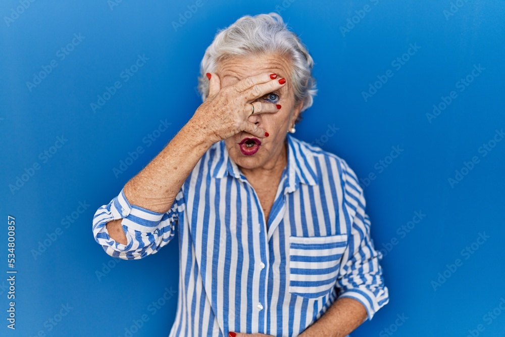 Senior woman with grey hair standing over blue background peeking in ...