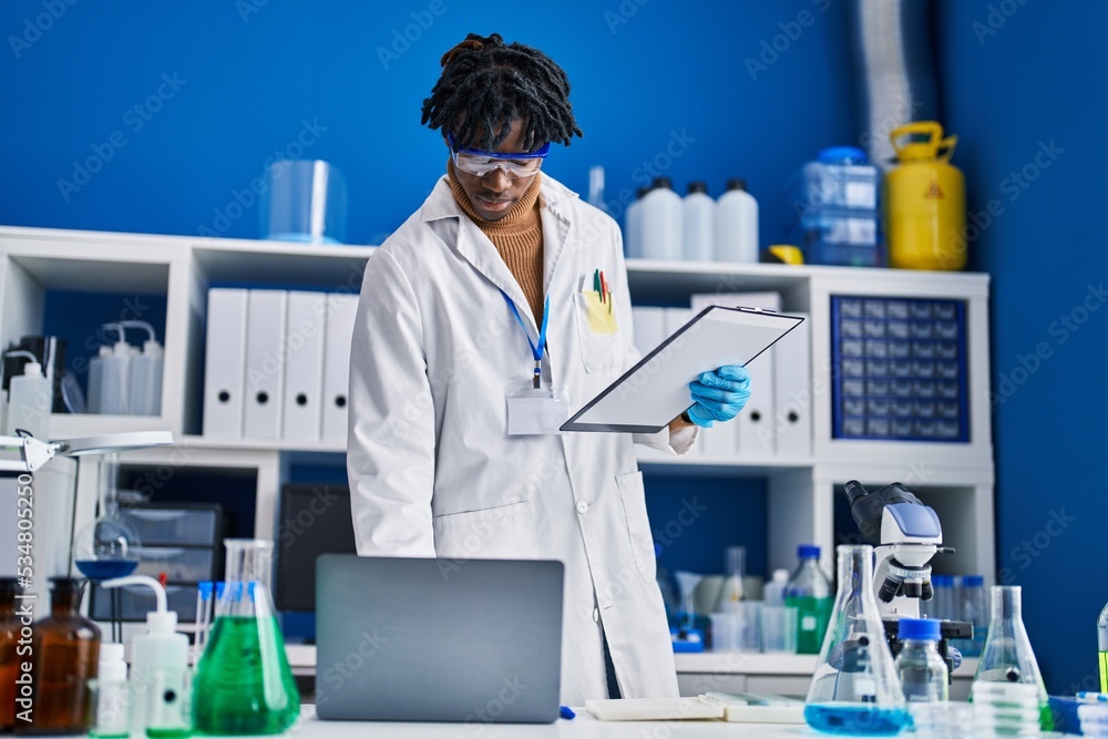 African american man scientist using laptop holding clipboard at ...