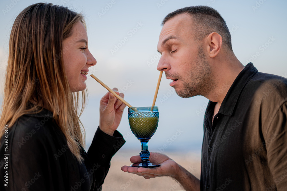 Young couple sitting on a beach, man accidentally gets drinking straw ...
