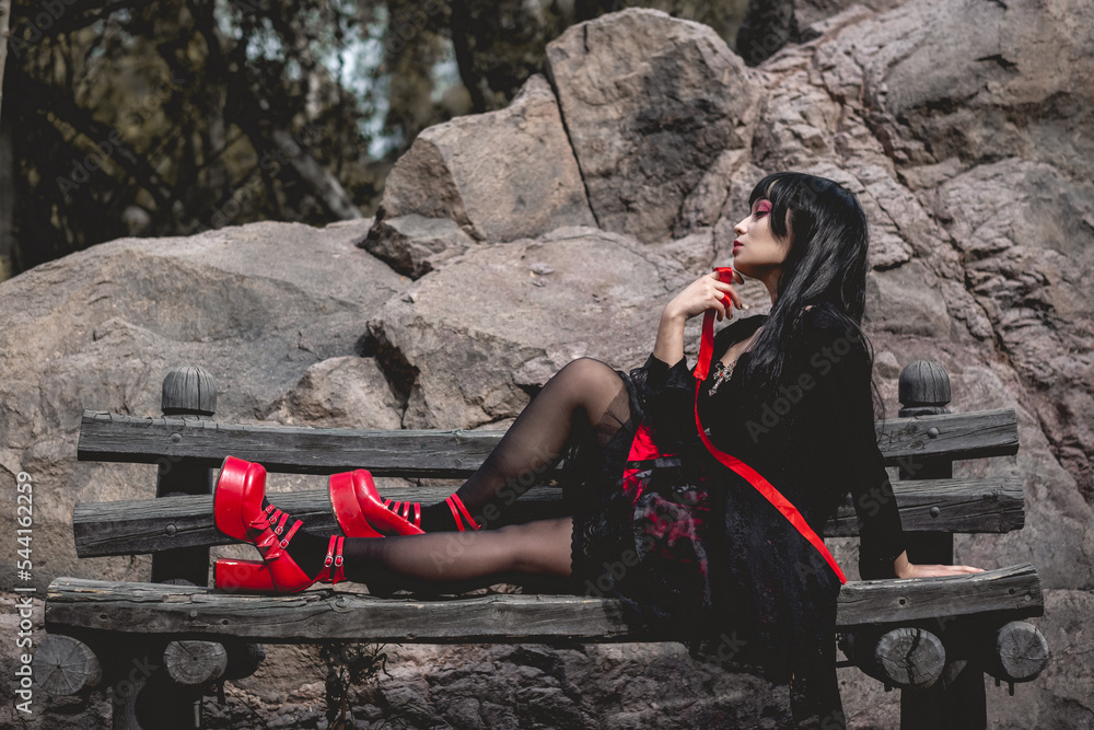 Young, sexy and vampiric hispanic goth girl with black dress and red platforms shoes seated and ...