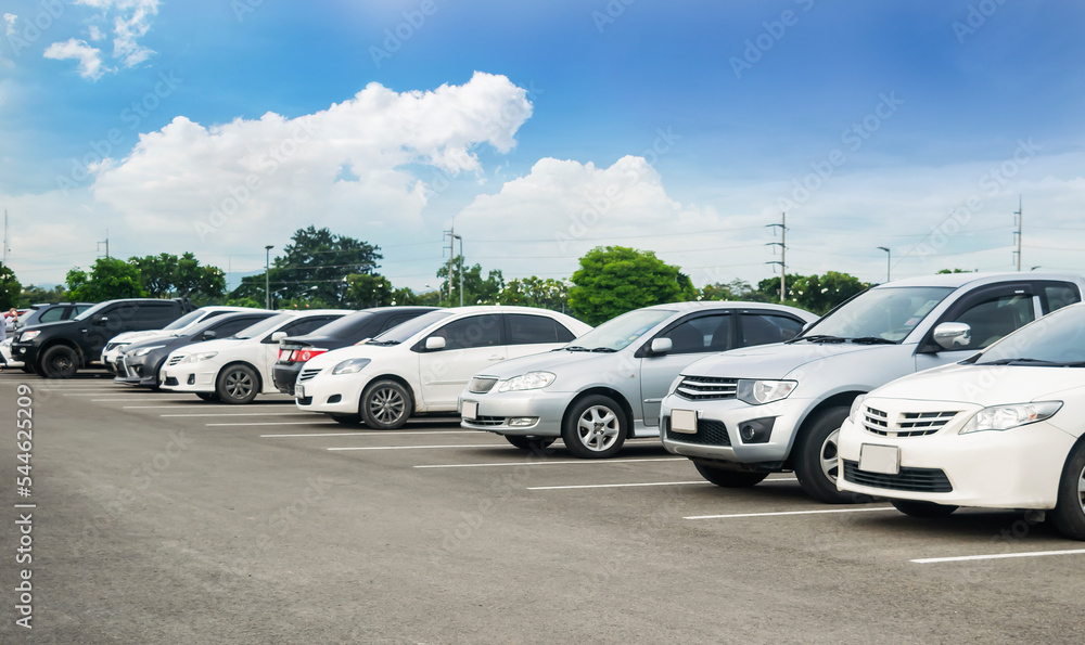Car parking in large asphalt parking lot with trees, white cloud and blue sky background ...