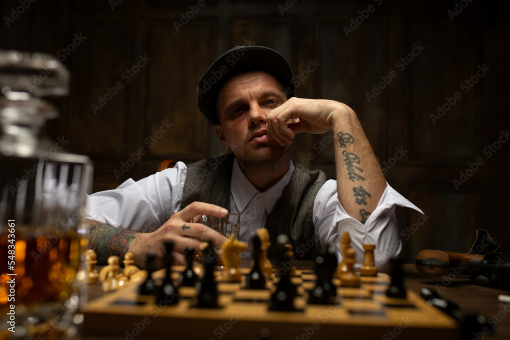 Young British gangster of twenties at table with chess and alcohol ...