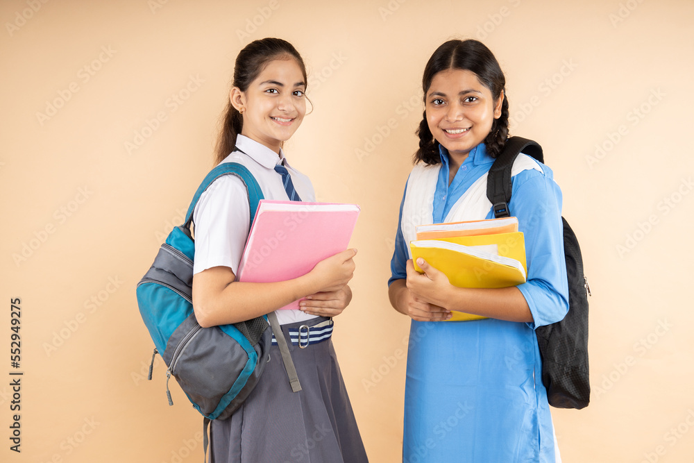 Happy Rural and Modern Indian student schoolgirls wearing school uniform holding books and bag ...