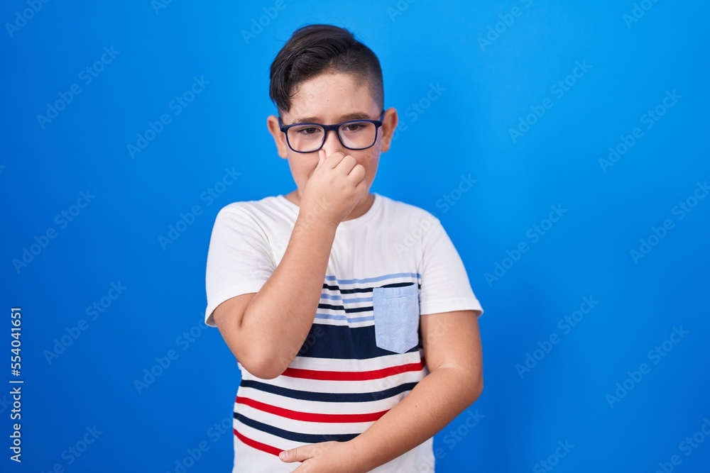 Young hispanic kid standing over blue background smelling something ...